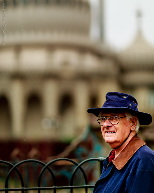 Portrait of an elderly man in a navy hat and blue coat against a rain-softened historic facade